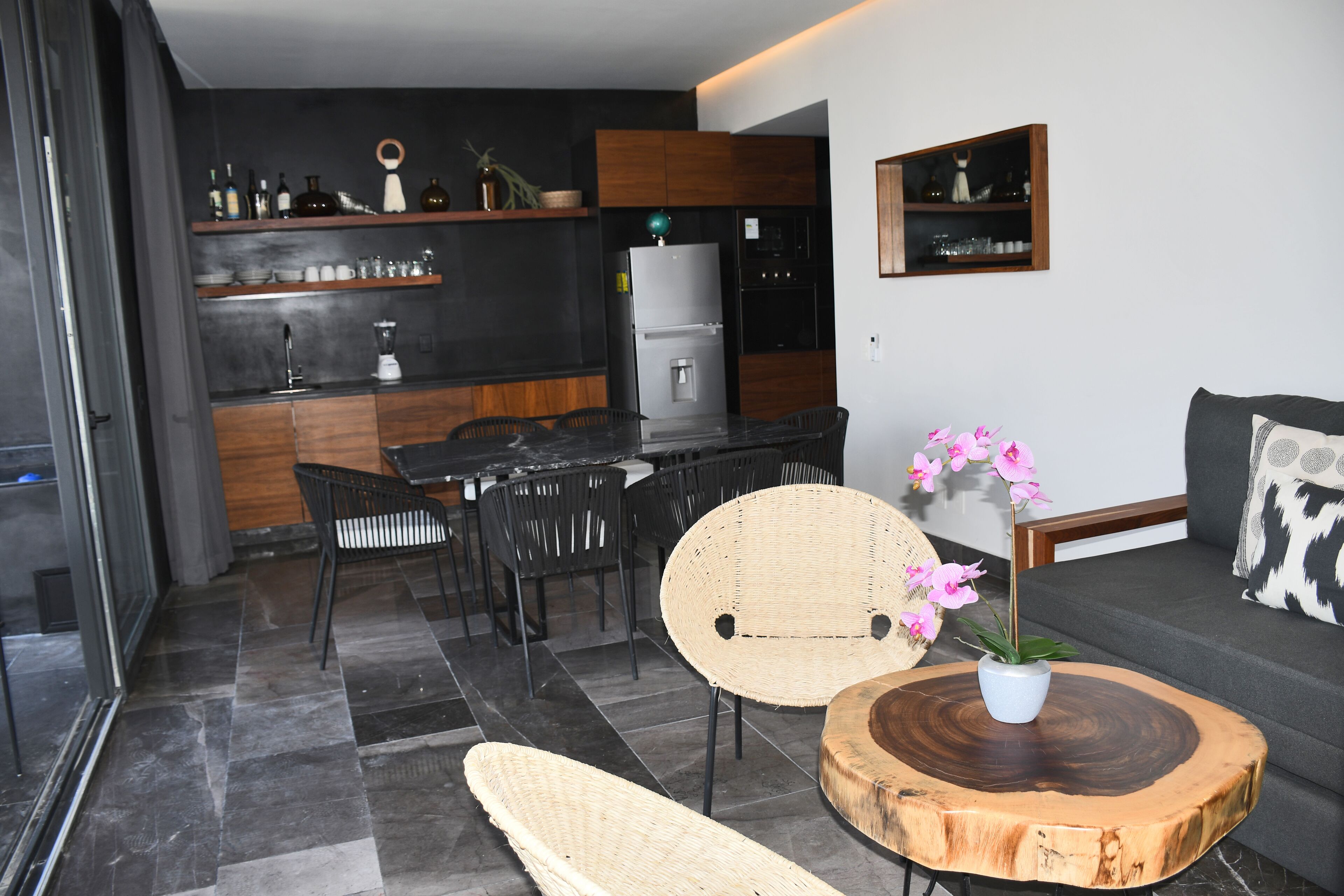 Uma cozinha moderna com armários escuros, uma sala de jantar com mesa redonda de madeira e uma sala de estar com sofá e almofadas decorativas.