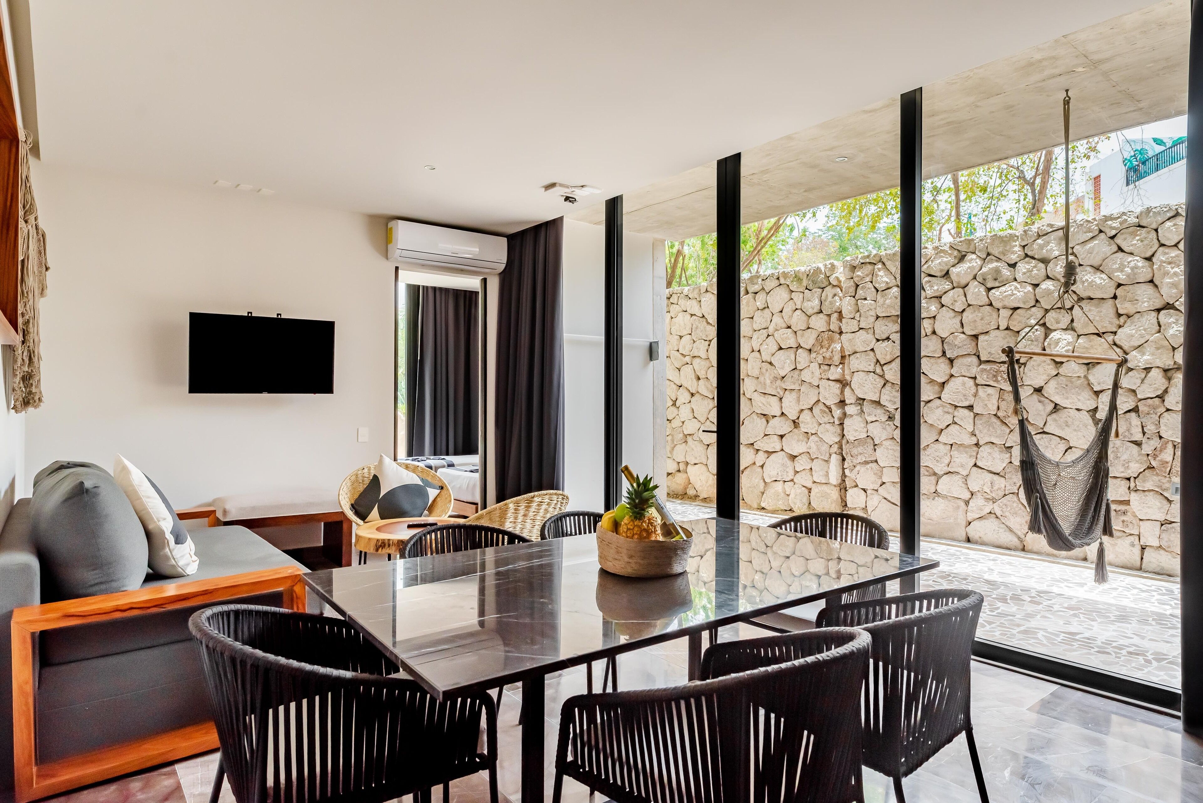 Uma área de jantar moderna com mesa de vidro, cadeiras pretas e parede de pedra ao fundo.