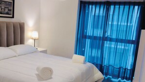 Quarto de hotel com cama, mesa de cabeceira, abajur e uma janela grande com cortinas azuis.