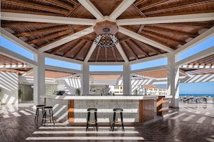 Uma área de bar ao ar livre espaçosa, com teto alto e vigas de madeira, destacando um balcão central com banquetes e vista para o oceano.