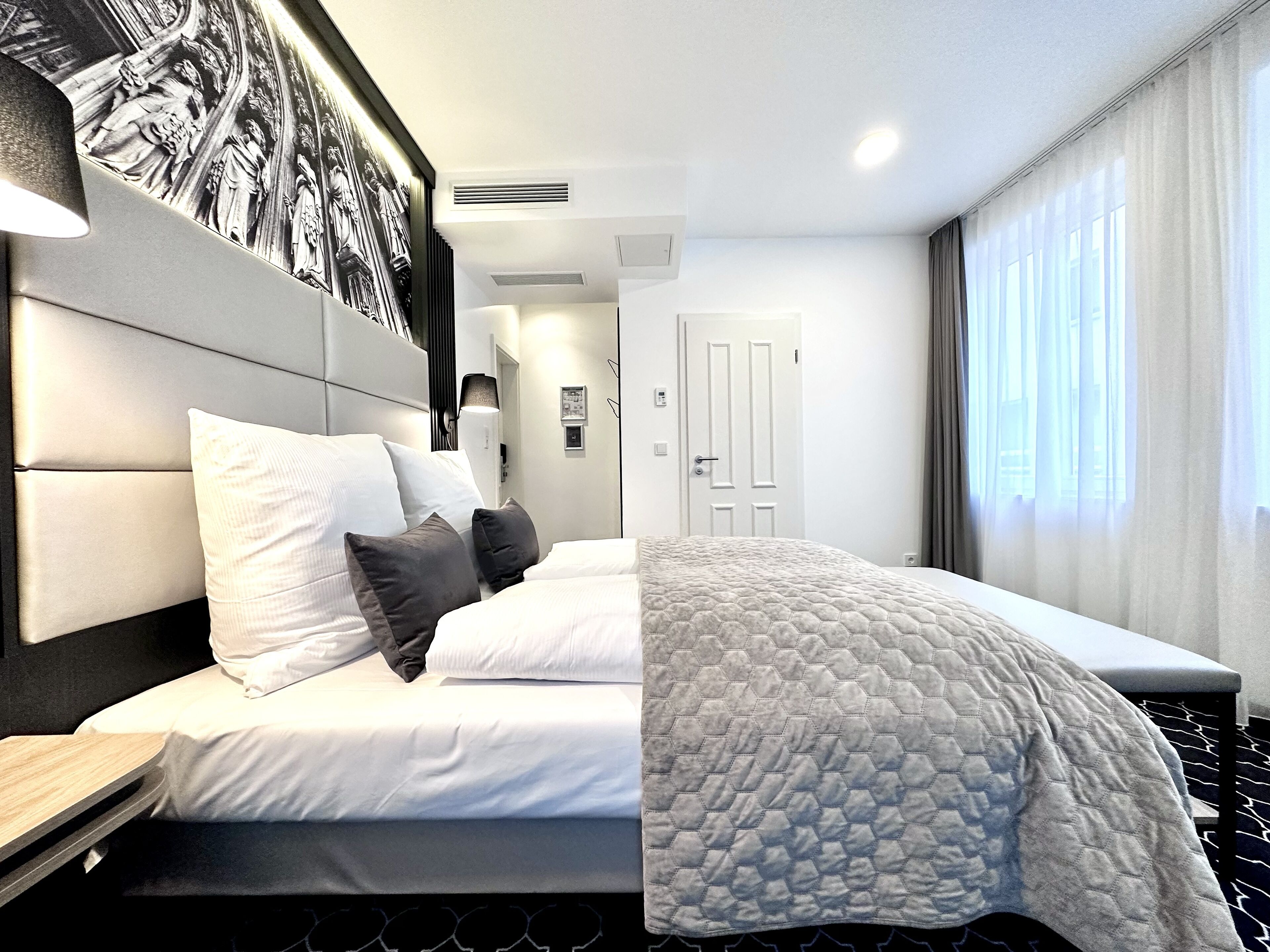 Quarto de hotel moderno com uma cama grande, cabeceira com um mural, mesa de cabeceira e vista para a paisagem urbana pela janela.