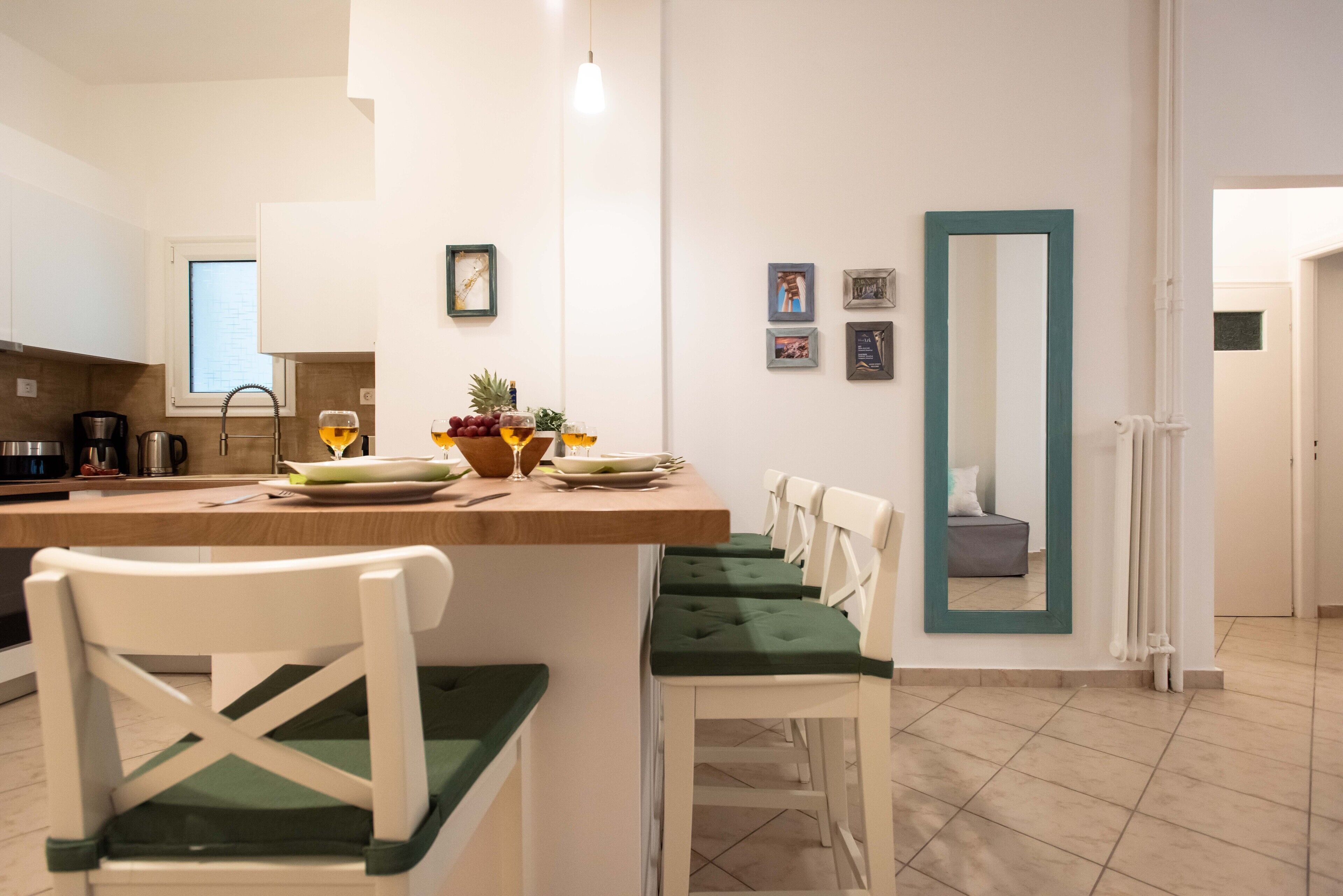 Cozinha moderna com armários brancos, bancada de madeira e área de refeições com cadeiras brancas e almofadas verdes. Há quadros nas paredes e um espelho com moldura azul-esverdeada.