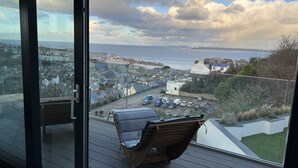 Uma varanda com uma cadeira de madeira, com vista para uma cidade costeira, carros estacionados e o mar.