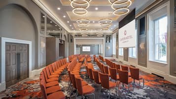 Uma sala de conferência com fileiras de cadeiras laranja, piso coberto de carpete e uma tela grande exibindo o logotipo do Clayton Hotel.