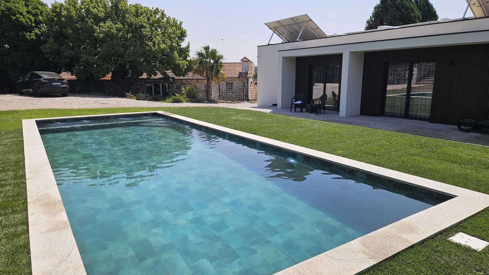 Piscina retangular com água cristalina, cercada por um gramado bem cuidado e um edifício moderno com amplas janelas.