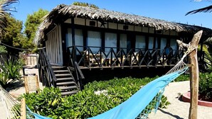 Uma casa de praia com telhado de palha, uma rede e uma escadaria que leva à entrada.