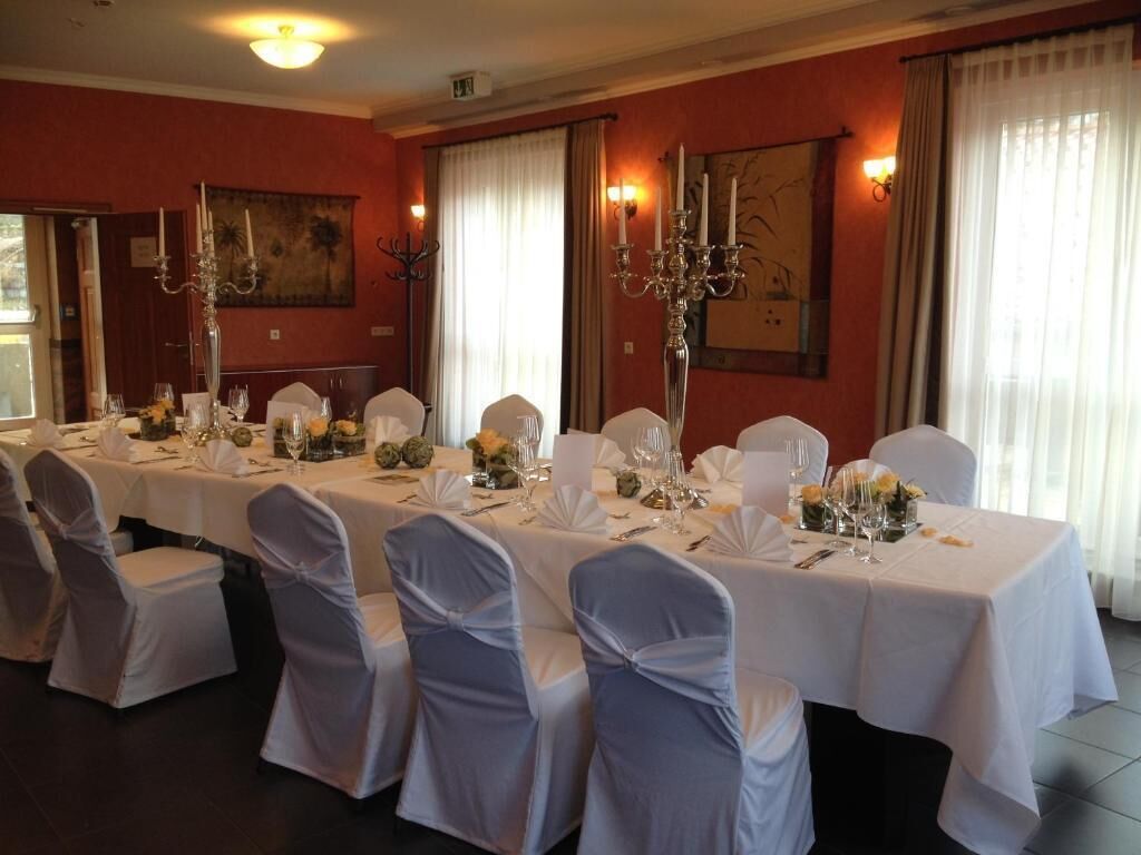 Uma sala de banquetes com mesas longas preparadas para um evento formal, cadeiras brancas e candelabros decorativos.