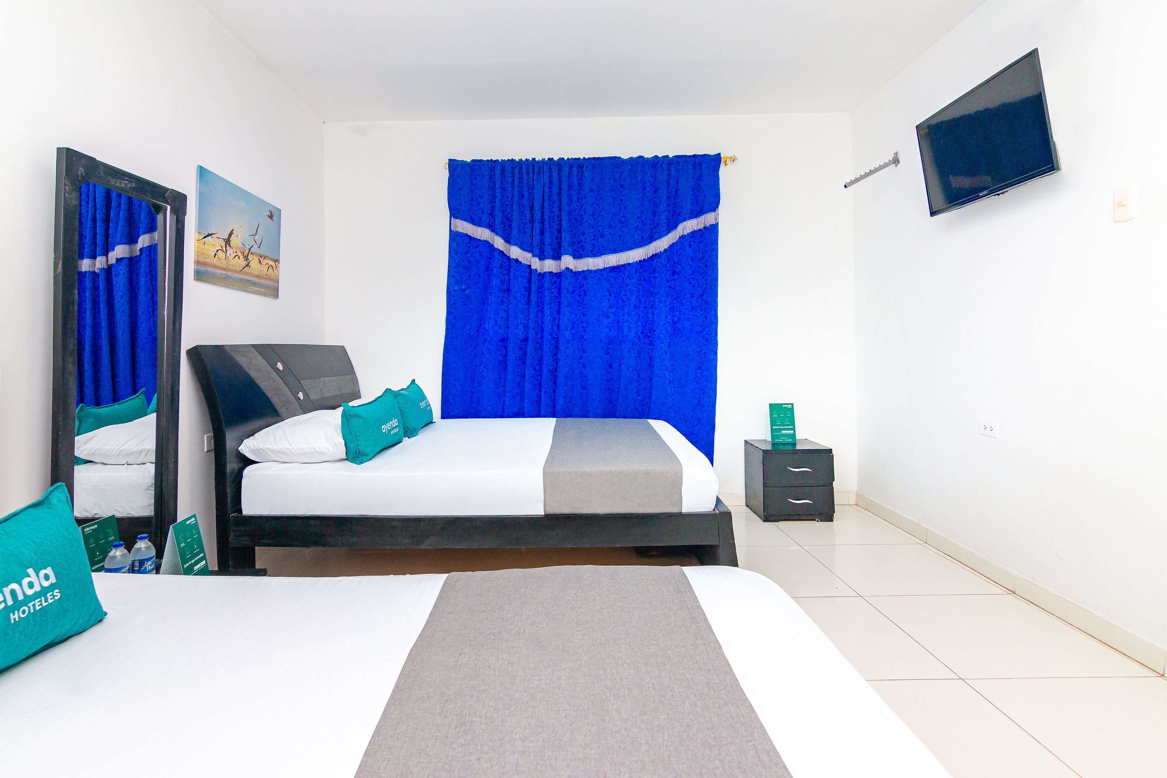 Quarto de hotel com uma cama, televisão de tela plana, janela com cortinas azuis e uma mesa de cabeceira pequena.