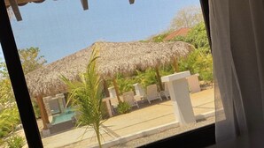 Vista pela janela de um resort, mostrando uma construção com telhado de palha, área de estar externa e piscina.