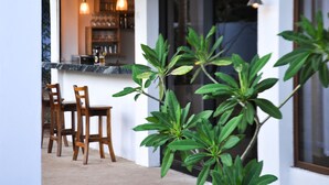 Uma área de pátio com cadeiras e mesa de madeira, um balcão com copos e garrafas, e plantas verdes.