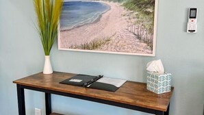 Quarto com escrivaninha, dois banquinhos, pintura de uma praia, ar condicionado e caixa de lenços.