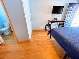 Quarto com cama, escrivaninha, cadeira, televisão e porta.