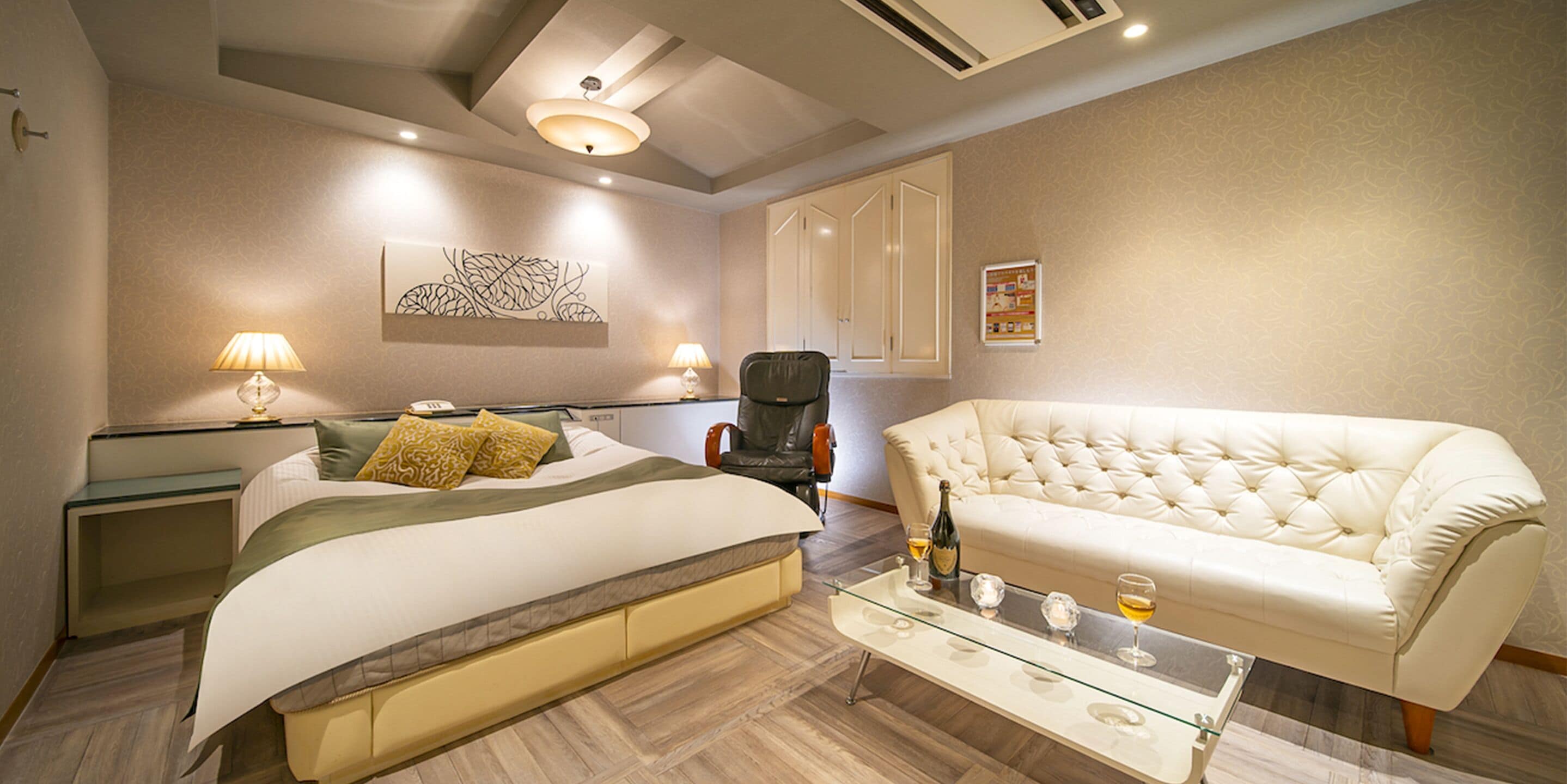 Quarto de hotel moderno com cama, mesa de centro de vidro com bebidas, sofá de couro e uma poltrona.