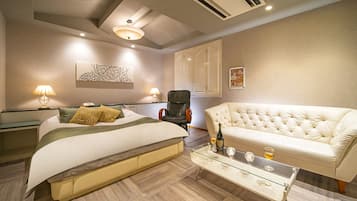 Quarto de hotel moderno com cama, mesa de centro de vidro com bebidas, sofá de couro e uma poltrona.