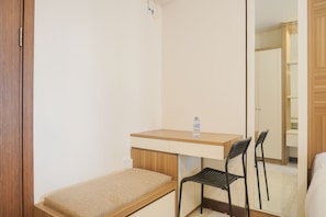 Quarto compacto com escrivaninha de madeira, um banco e uma cadeira. Uma garrafa de água está sobre a escrivaninha.