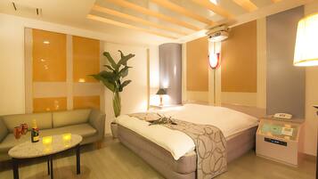 Quarto de hotel com uma cama, um sofá, uma pequena mesa com bebidas, uma planta, um abajur e um painel de controle.