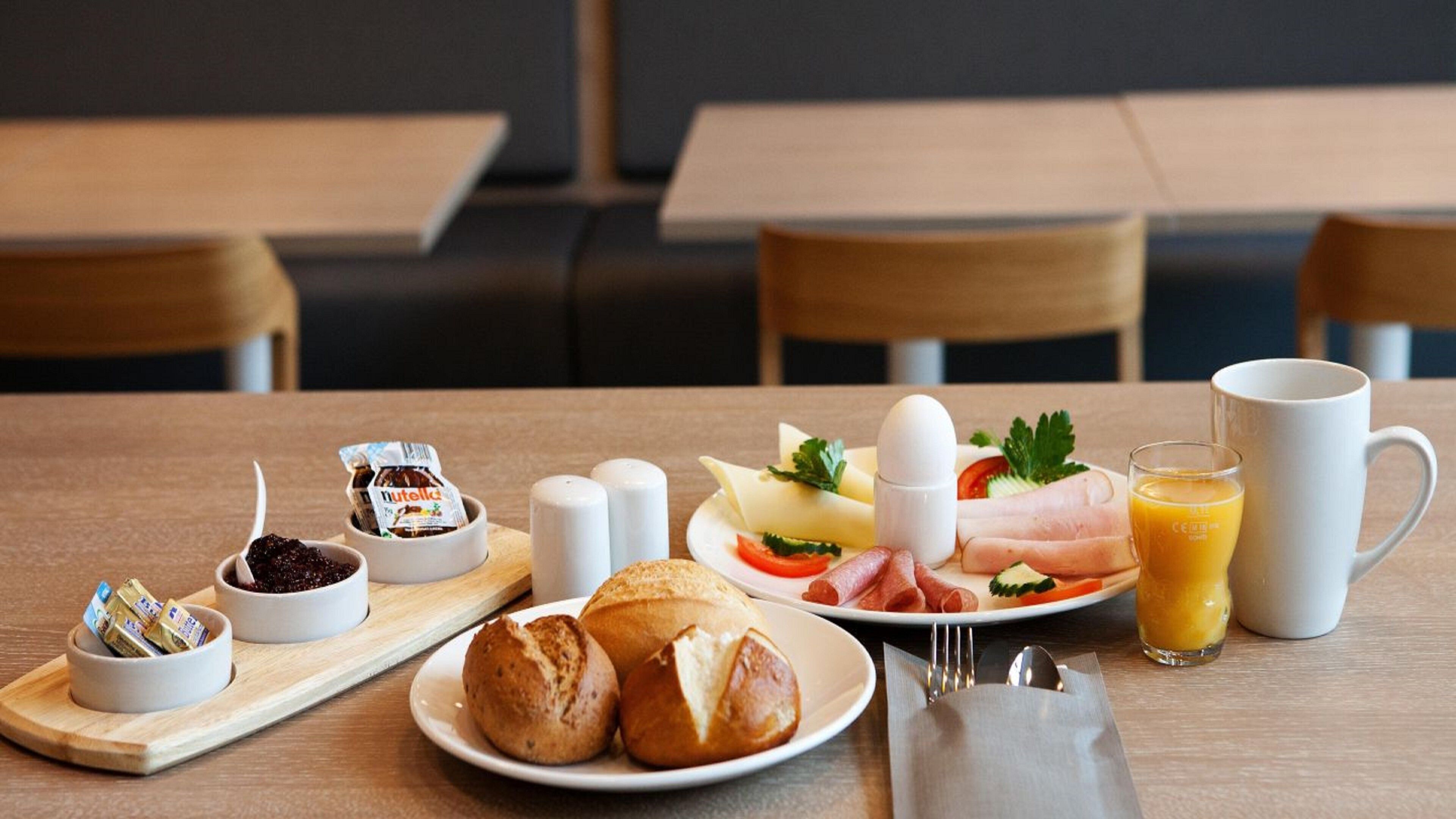 Uma mesa de café da manhã com um prato de ovos, presunto, legumes e pães, acompanhados de manteiga, geleia e suco de laranja.