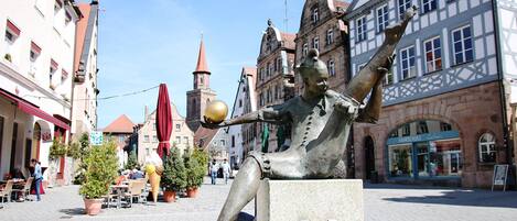 Uma estátua de um homem com espada e escudo em uma praça pública com calçamento de pedra e edifícios históricos ao fundo.