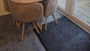 Uma área de refeições com uma mesa de madeira redonda, duas cadeiras estofadas, um relógio de parede decorativo e vista para uma área externa gramada através de uma porta de vidro.