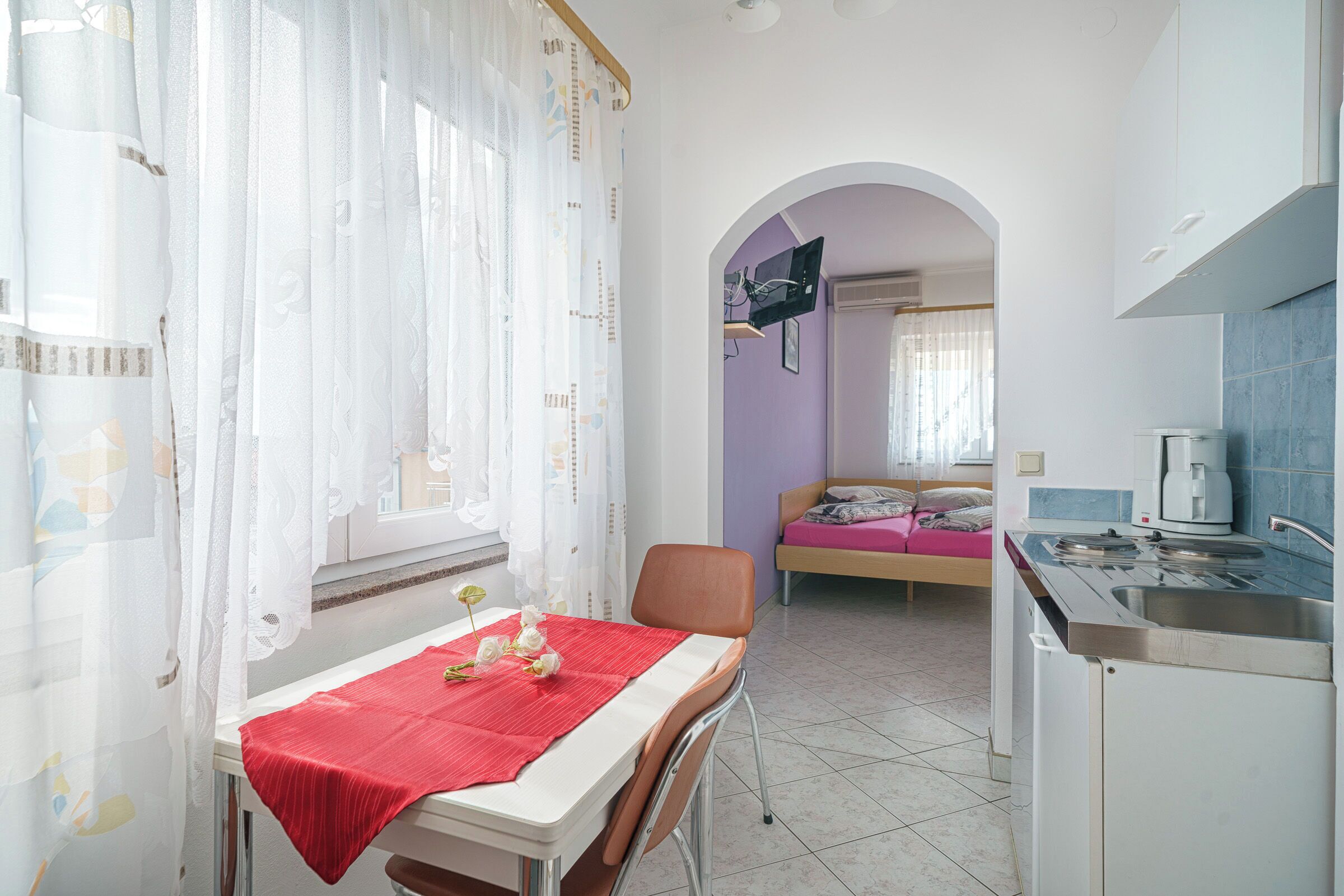 Quarto de hotel compacto com mesa de jantar, cozinha compacta, cama e televisão fixada na parede.