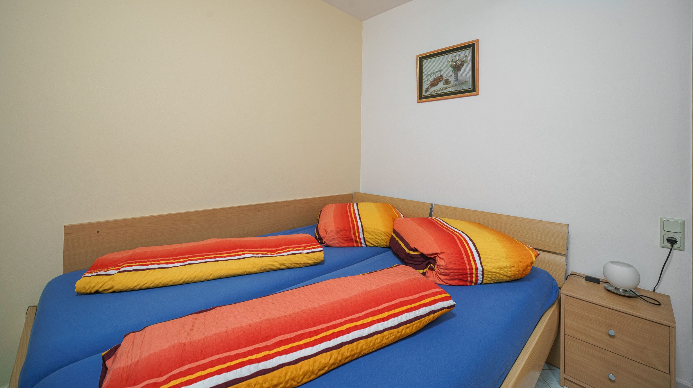 Uma cama com lençóis azuis e almofadas listradas e coloridas, uma pequena mesa de cabeceira com uma luminária e um quadro na parede.