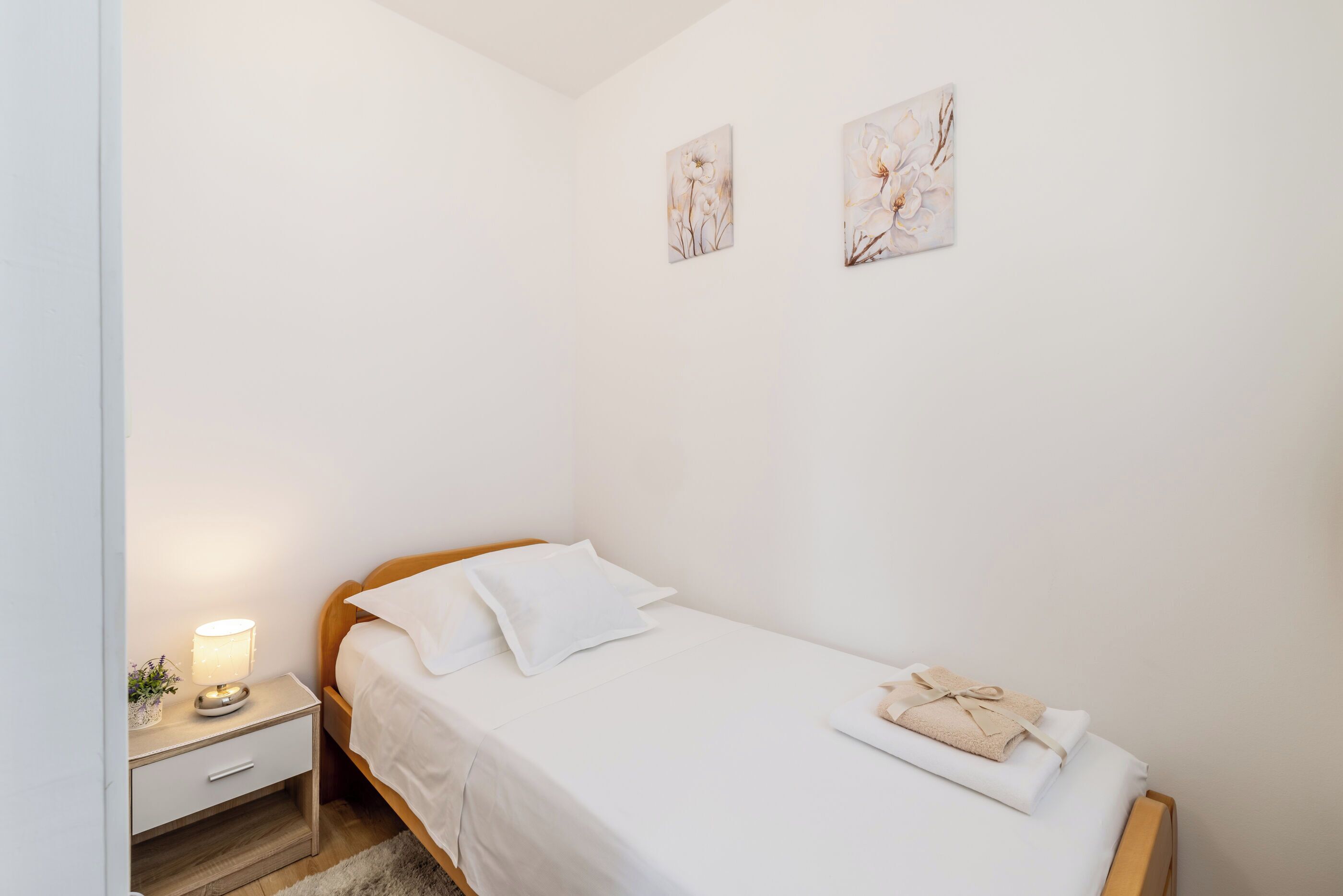 Quarto bem arrumado com cama de madeira, roupa de cama branca, abajur e duas obras de arte floral nas paredes.