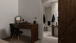 Quarto moderno e minimalista, com escrivaninha de madeira, cadeira preta e espelho. Porta de madeira e pequena mesa com garrafas e uma vela.