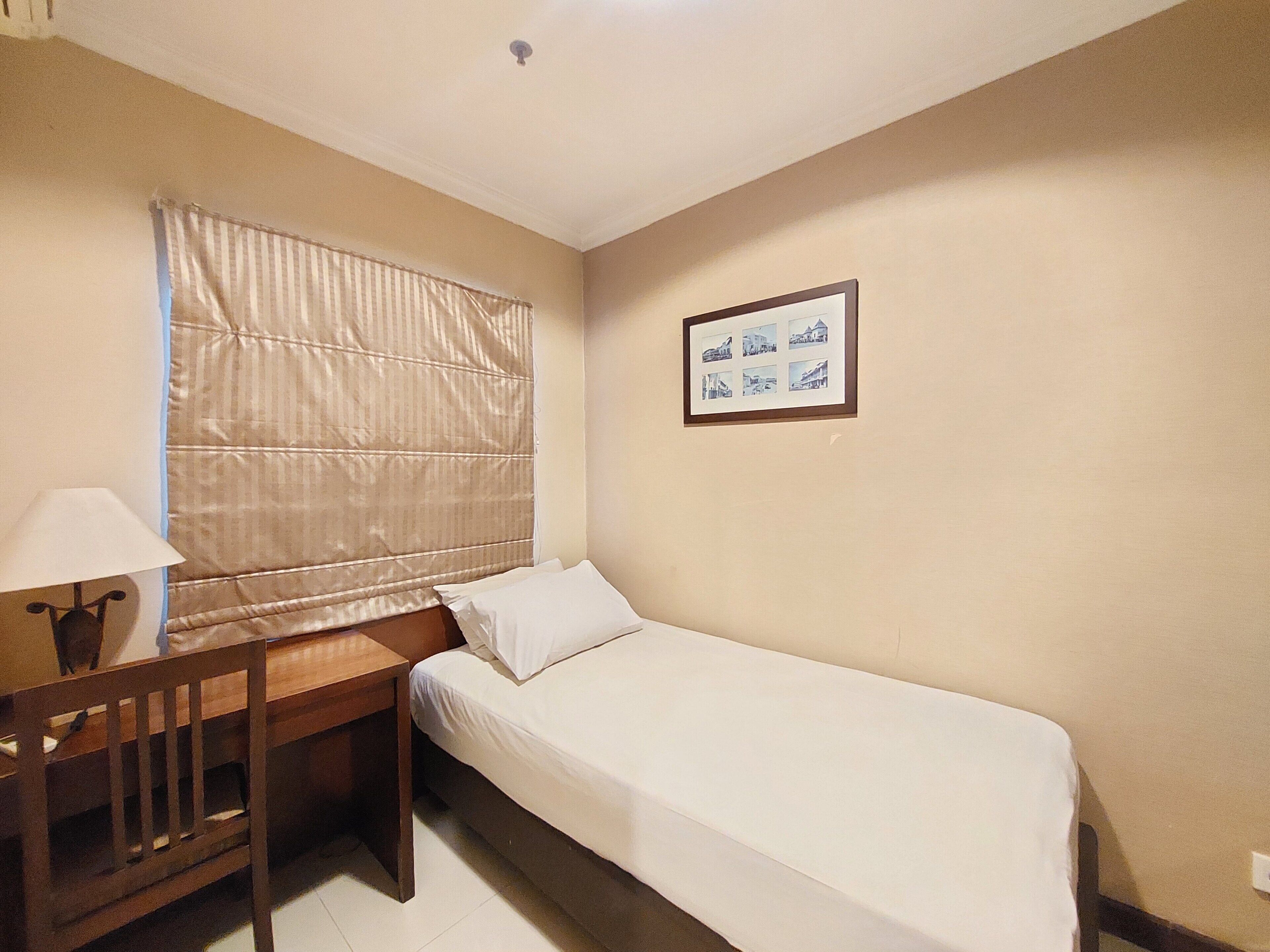 Quarto pequeno e bem arrumado, com uma cama de casal, uma escrivaninha e uma cadeira. Há uma janela com cortina e um quadro na parede.
