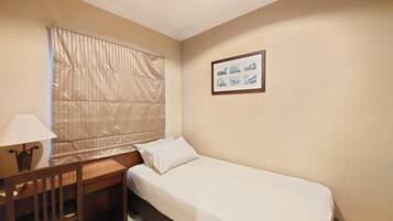 Quarto pequeno e bem arrumado, com uma cama de casal, uma escrivaninha e uma cadeira. Há uma janela com cortina e um quadro na parede.