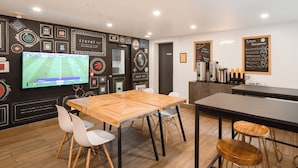 Uma sala de reuniões moderna com uma mesa grande, cadeiras brancas, um espaço de bar e uma televisão exibindo um jogo esportivo.