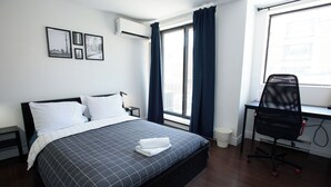 Cama bem arrumada com colcha xadrez, uma escrivaninha com cadeira e uma janela com cortinas.