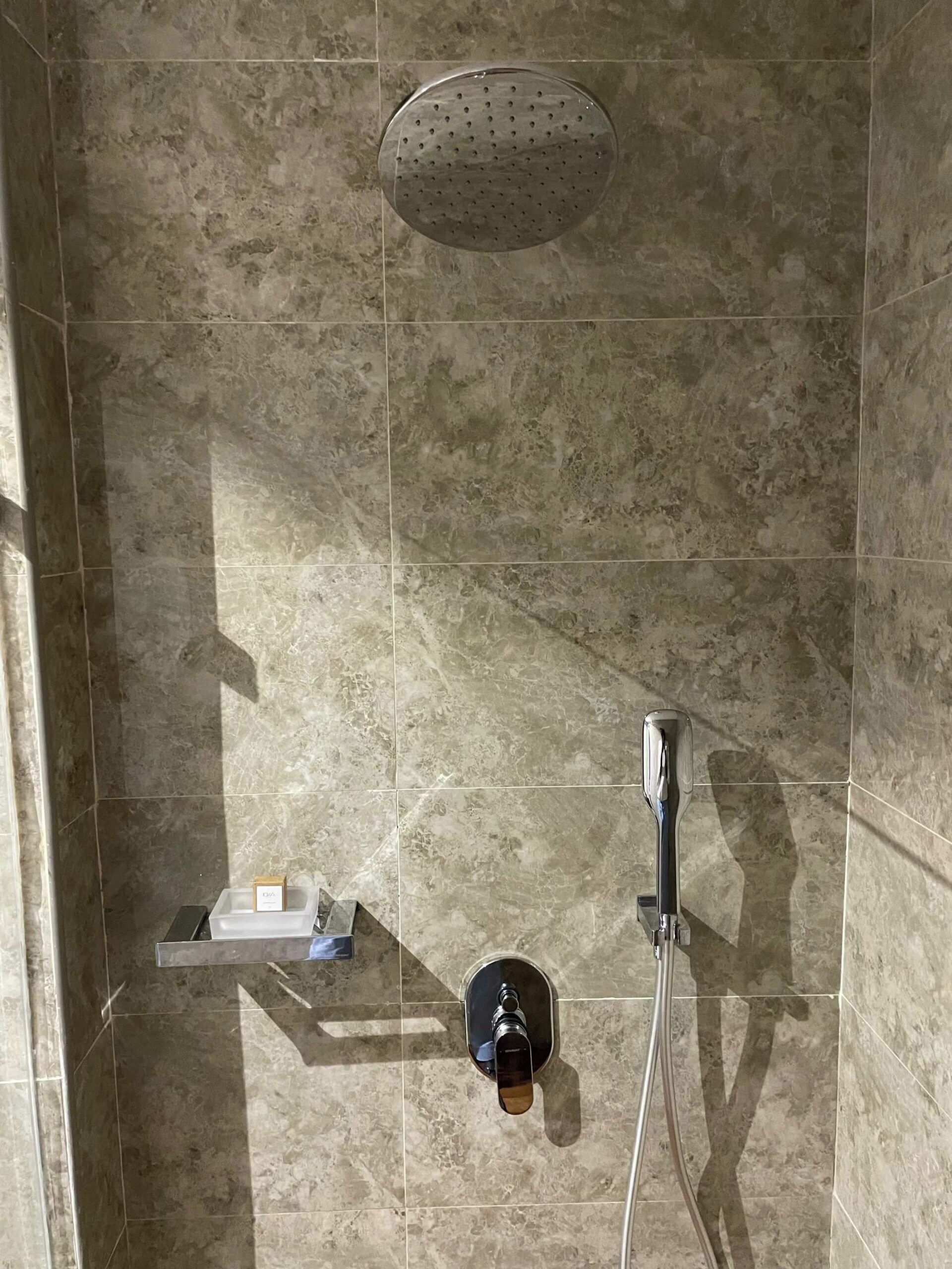 Área de chuveiro com chuveiro manual, painel de controle fixado na parede e piso revestido com azulejos.