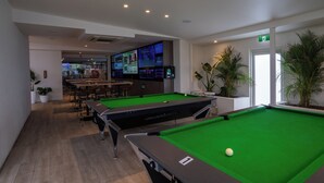 Uma mesa de bilhar com superfície de feltro verde, uma taça de bilhar e uma bola branca. Várias mesas e cadeiras no fundo, e um bar com uma tela de televisão exibindo diversos canais.
