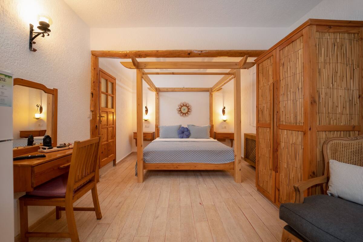 Quarto com cama de madeira, uma escrivaninha e uma cadeira. Há um aplique na parede e um relógio de parede.