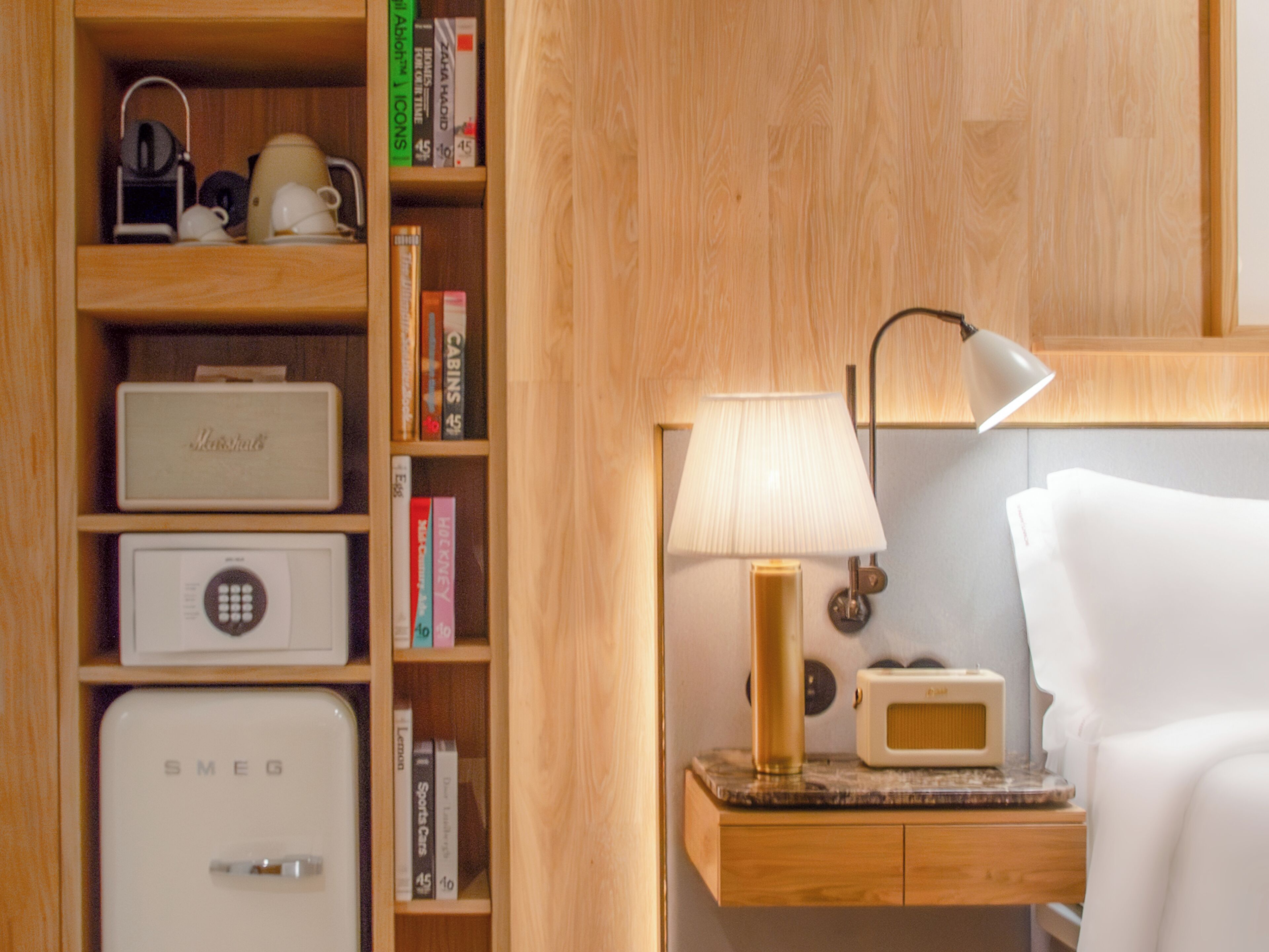 Quarto de hotel com mesa de cabeceira de madeira, luminária, rádio e estante com vários livros e um despertador antigo.