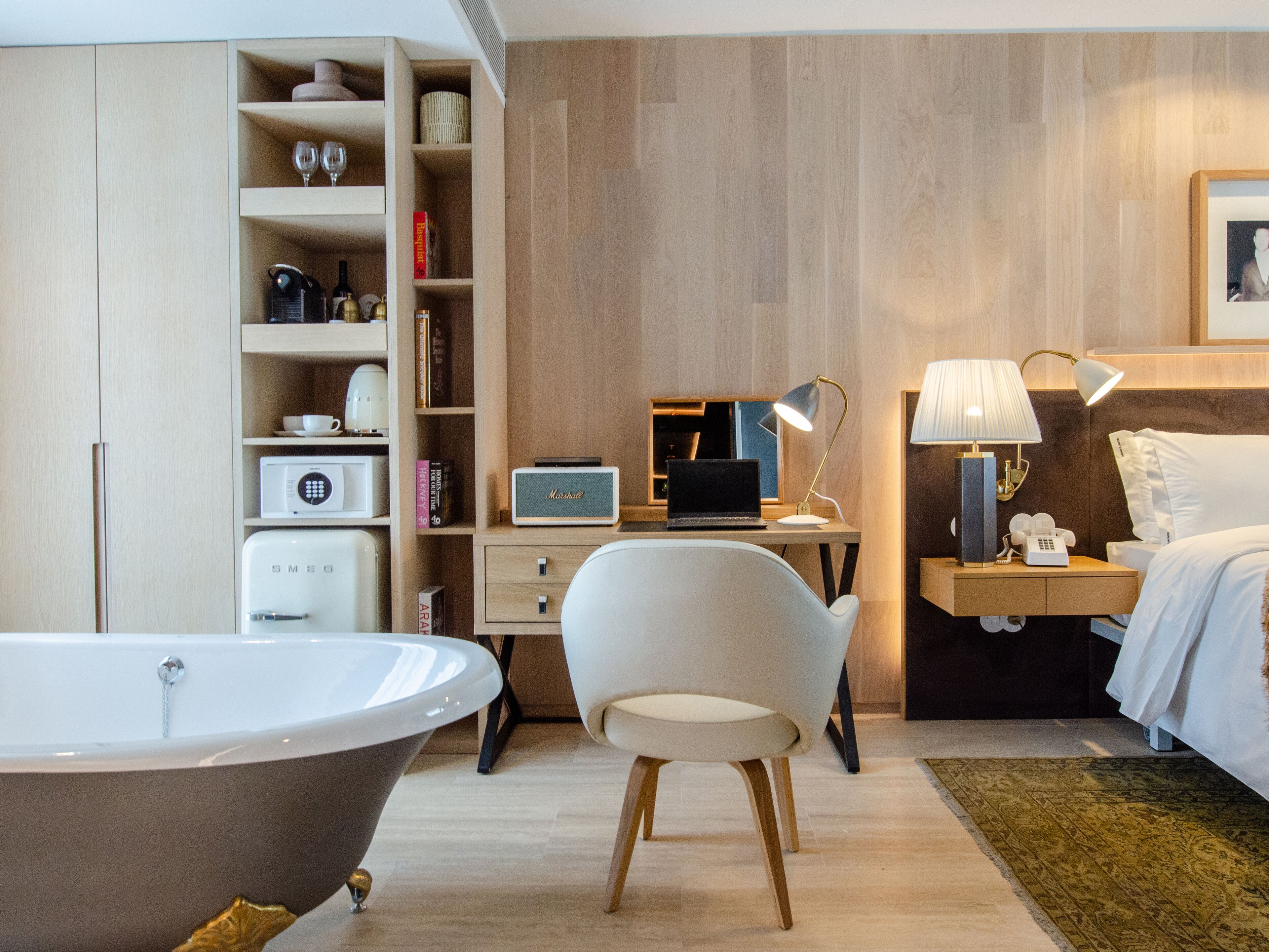 Quarto moderno com banheira, escrivaninha com laptop, mesa de cabeceira com abajur e estante de livros.