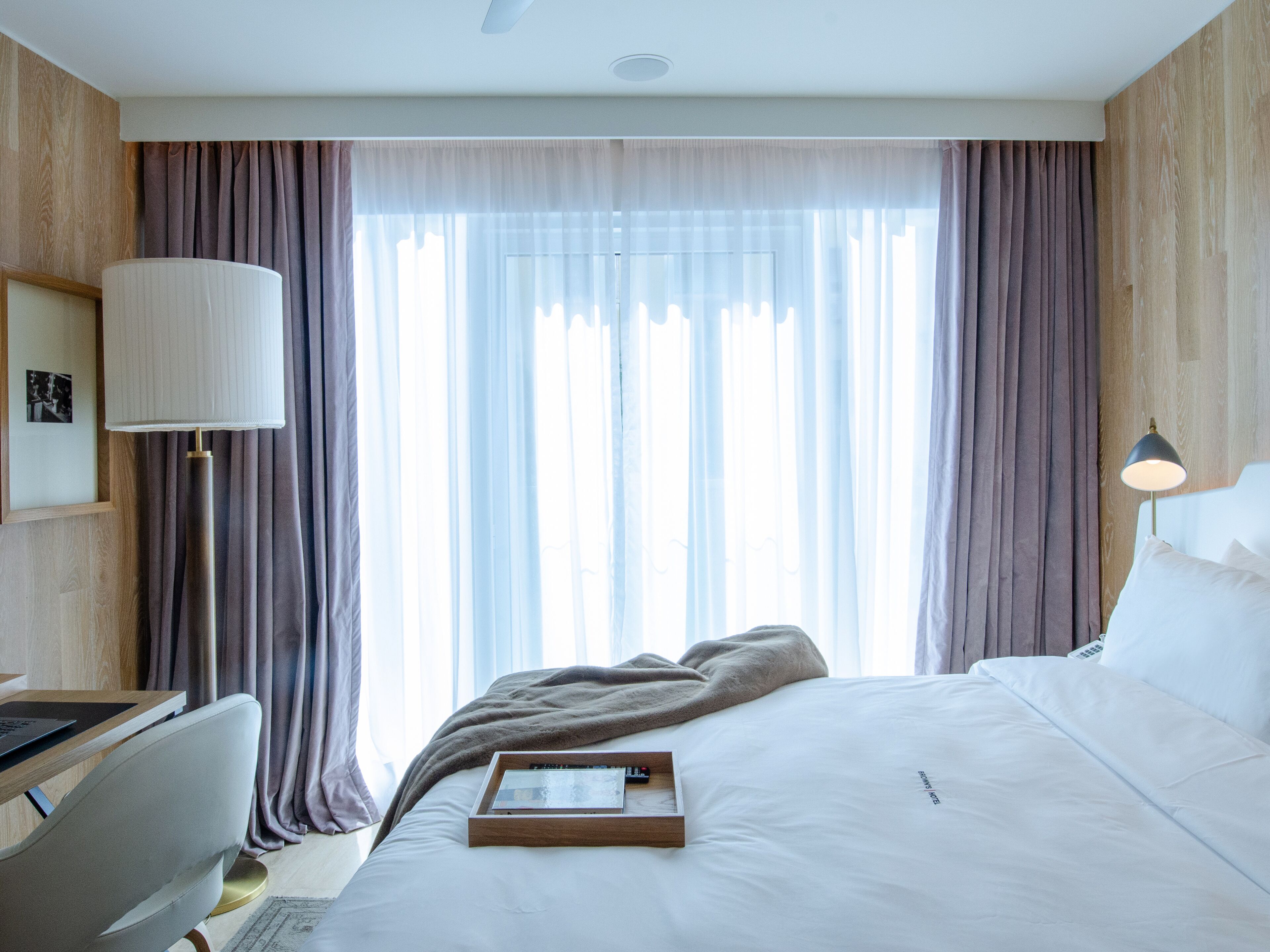 Quarto de hotel com cama, escrivaninha, luminária e janela com cortinas.