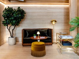 Uma sala de estar moderna com um sofá de veludo escuro, uma mesa de centro redonda de madeira, um abajur e uma pequena mesa lateral com livros.