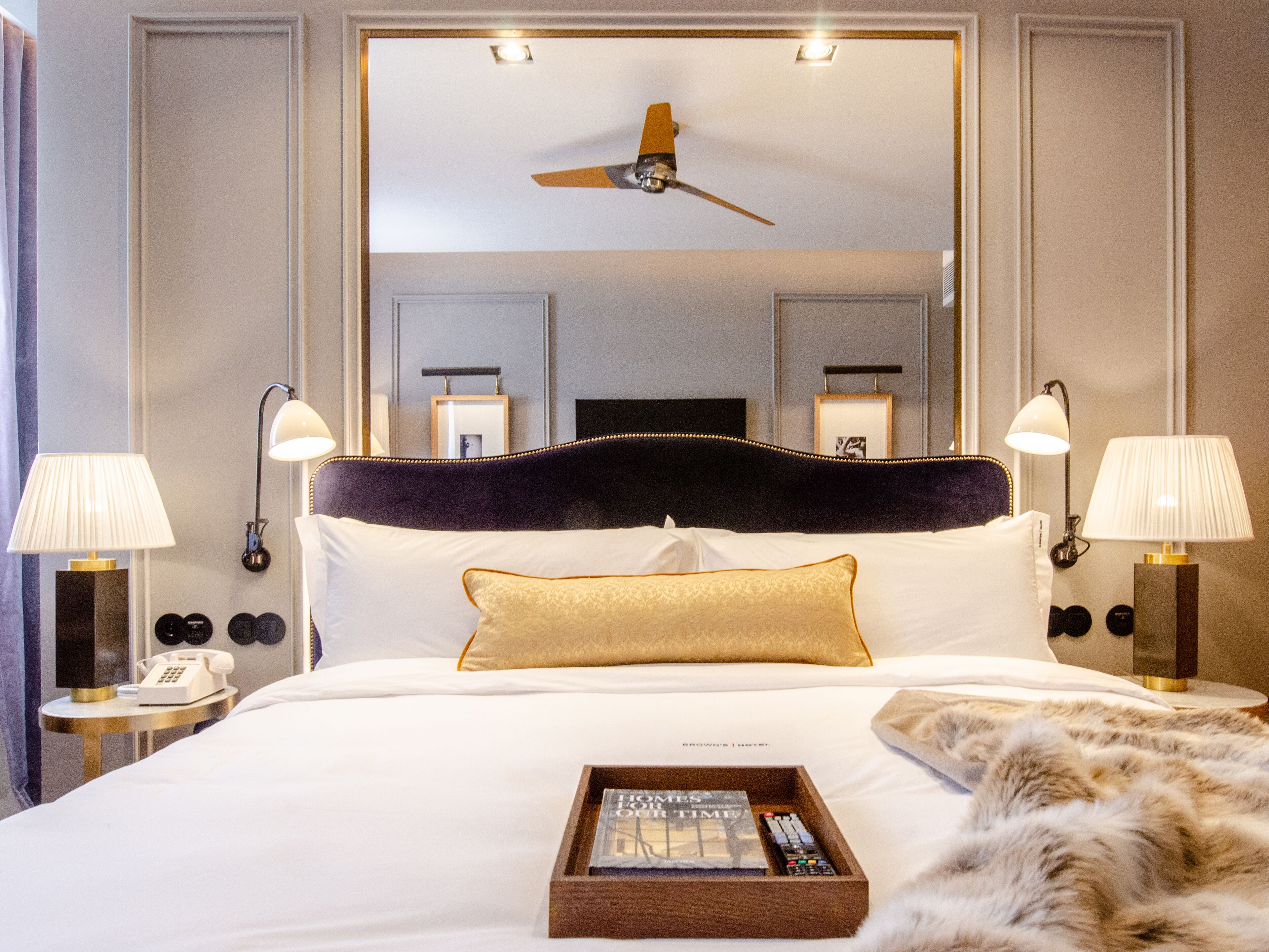 Quarto de hotel com uma cama, duas luminárias laterais, um ventilador de teto, telefone ao lado da cama e um bandejas com um controle remoto e um livro.