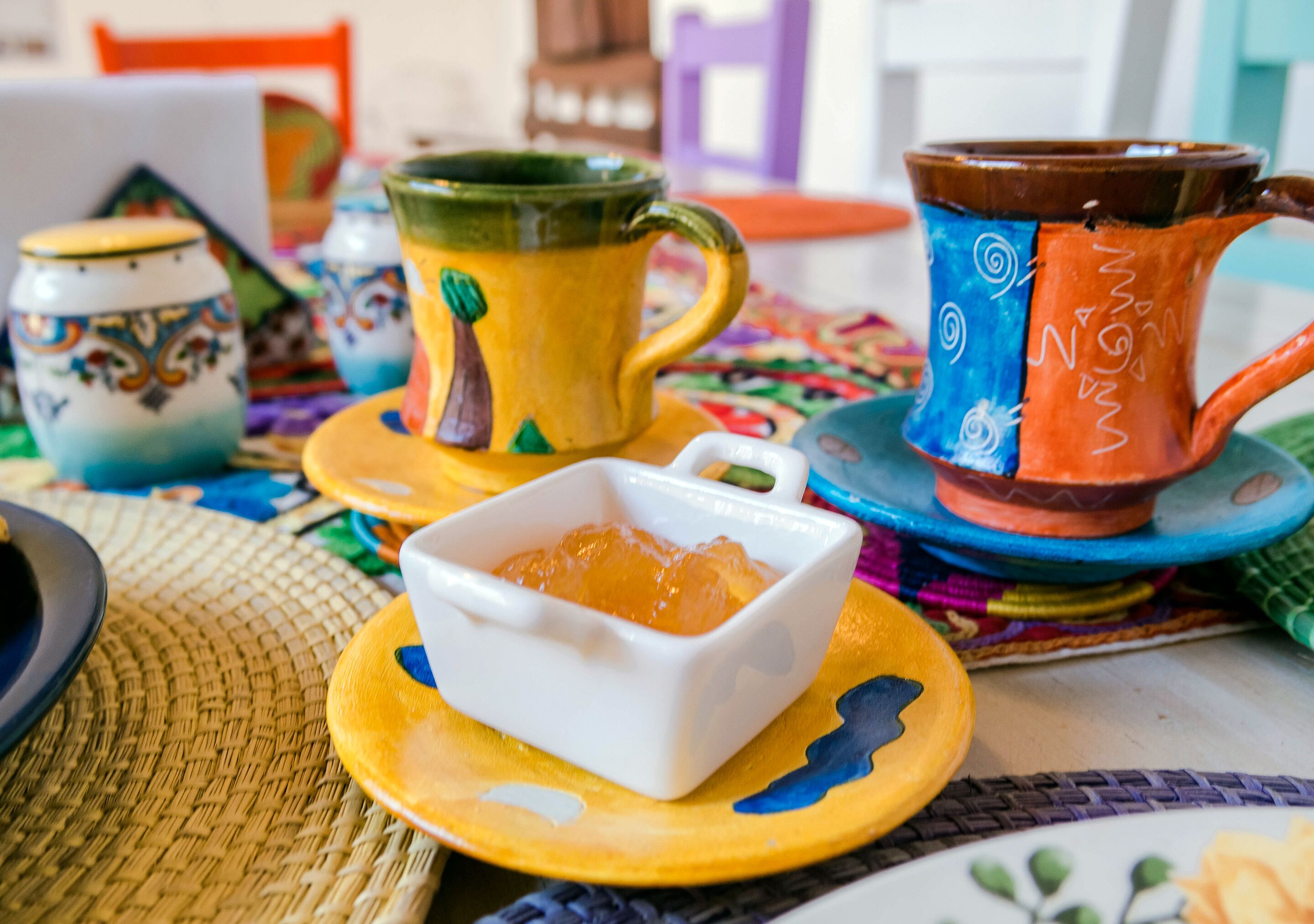 Uma mesa com um jogo americano trançado, uma tigela branca com um alimento amarelo, e xícaras e pratos de cerâmica colorida.