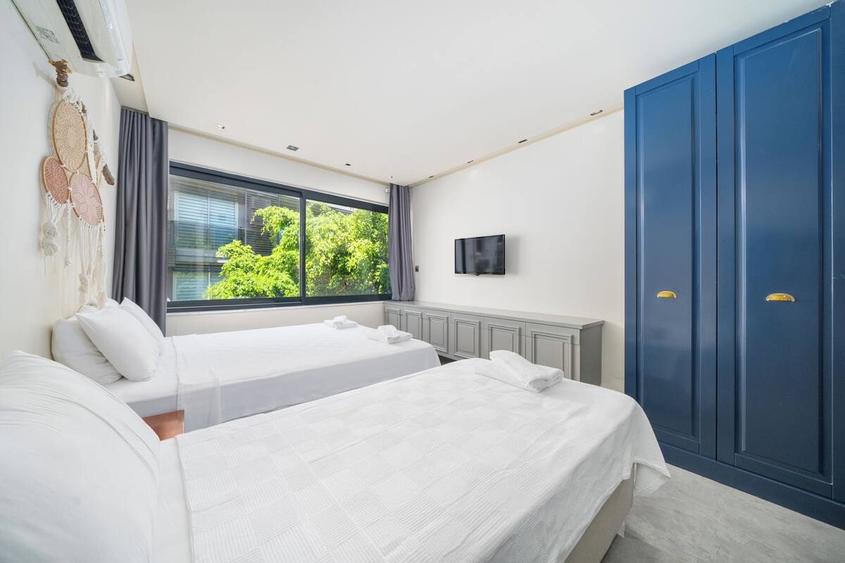 Quarto com duas camas, uma janela grande com vista para áreas verdes, um guarda-roupa azul e uma televisão de tela plana na parede.