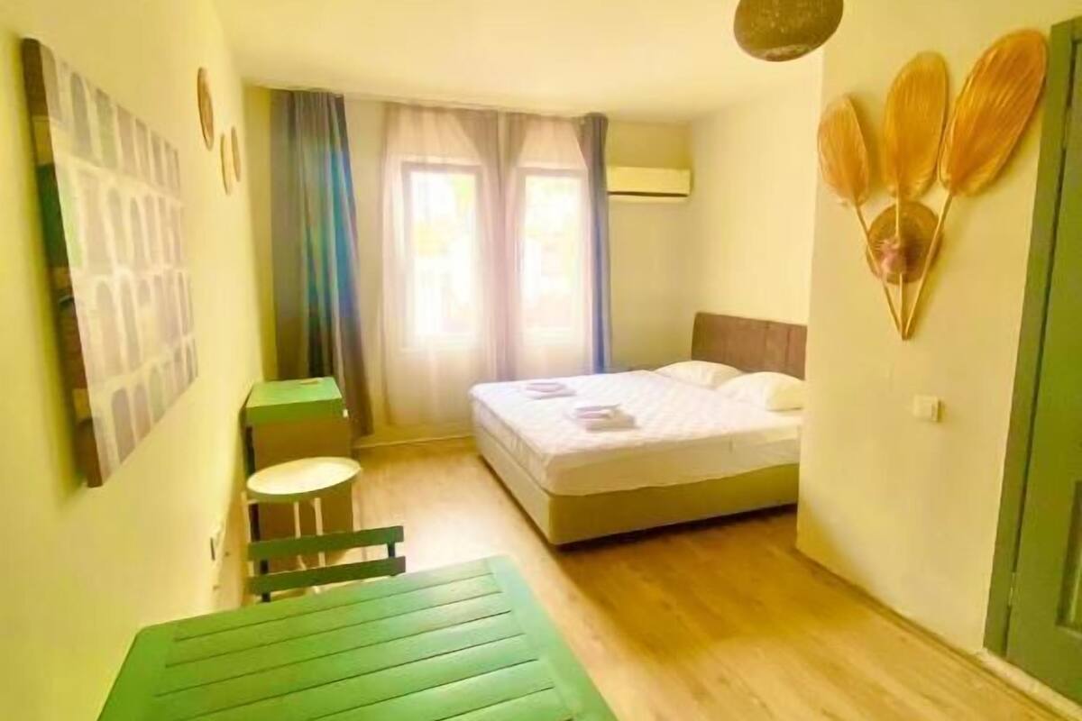 Quarto com cama, mesa e cadeira. Tem uma janela com cortinas, ar condicionado fixado na parede e um enfeite de parede.