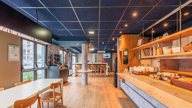 Interior de um café moderno com mesas de madeira, um balcão com alimentos e uma tela de TV ao fundo.