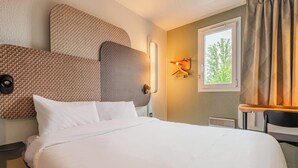 Uma cama bem arrumada com roupa de cama branca, luminária fixada na parede e uma janela com cortinas.