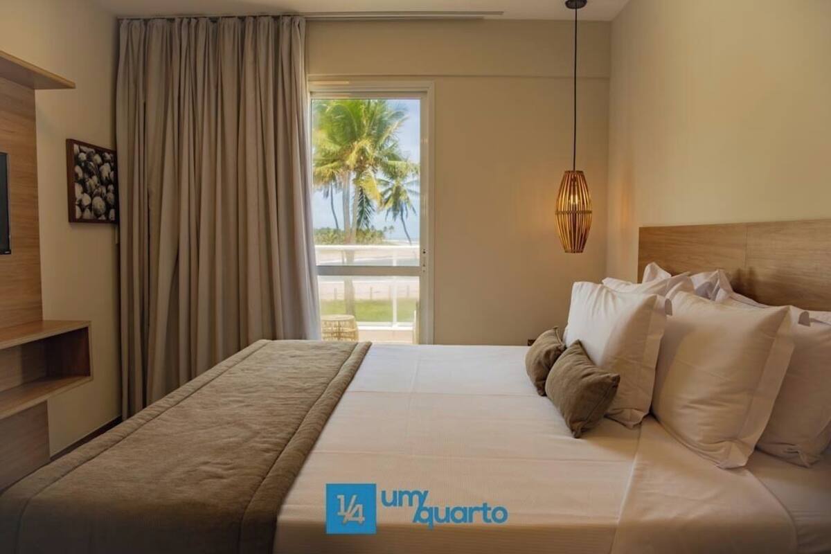 Quarto de hotel com cama, mesa de cabeceira, luminária pendente e vista para palmeiras pela janela.