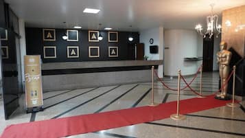 Uma área de recepção de hotel com um tapete vermelho que leva ao balcão, quadros nas paredes e uma estátua dourada.