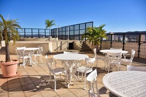 Um pátio externo com mesas e cadeiras de metal branco, plantas em vasos e vista para o mar.