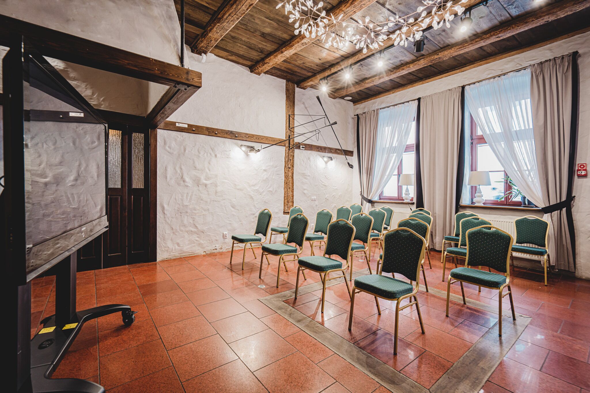 Sala de conferências com teto de madeira, paredes brancas e chão em azulejos. Há fileiras de cadeiras verdes e uma tela de projeção.