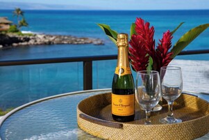 Uma garrafa de champanhe Veuve Clicquot Brut, dois copos e uma flor vermelha sobre uma bandeja de palha, com vista para o mar.