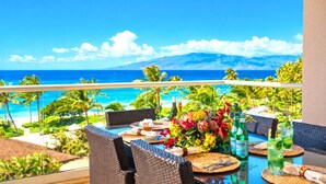 Uma mesa de jantar posta com cadeiras de vime, um arranjo floral e bebidas em uma varanda com vista para uma praia, palmeiras e uma montanha ao fundo.